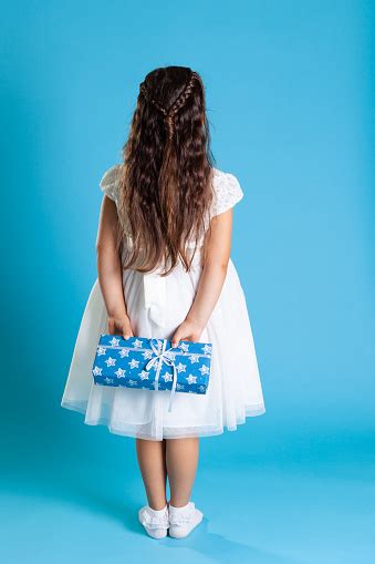 흰색 드레스를 입은 전체 길이의 작은 공주와 그녀의 등과 함께 서서 파란색 배경에 고립 된 크리스마스 또는 생일 선물을 숨깁니다 갈색 머리에 대한 스톡 사진 및 기타 이미지