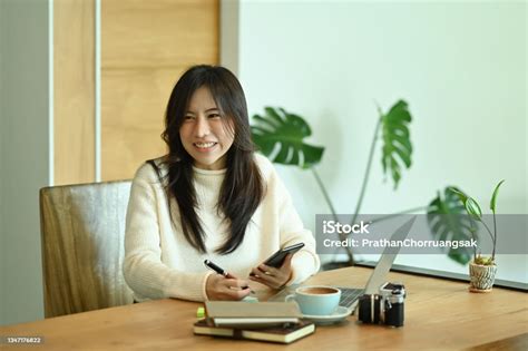 휴대 전화를 들고 사무실에서 컴퓨터 노트북으로 작업 하는 쾌활한 젊은 여자 25 29세에 대한 스톡 사진 및 기타 이미지 25 29세 30 34세 Brand Name