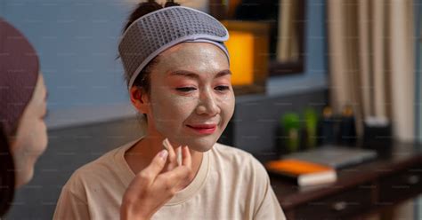 웃는 아시아 여자 친구들은 침대에 앉아 집에서 함께 얼굴에 스킨 케어 페이셜 마스크를 바르고 있다 여성 게이 커플은 긴장을