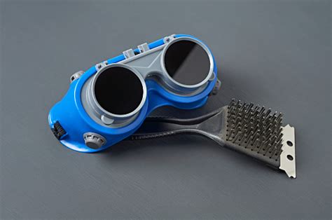 플라스틱 손잡이가있는 금속 브러시 근처에서 용접 할 때 눈을 보호하기위한 이중 유리가있는 파란색 고글은 작업장에서 어두운 콘크리트 책상에 놓여 있습니다 클로즈업 개성 개념에