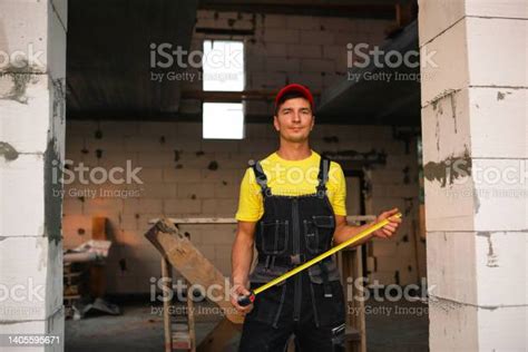 건설 현장의 건설 노동자 엔지니어는 줄자로 창문 개구부와 벽돌 벽의 길이를 측정합니다 작업복을 입은 남자 점프 슈트와 모자