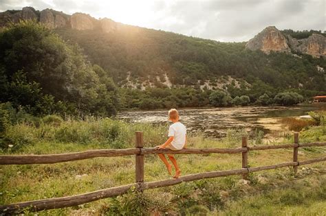 카메라에 등을 대고 있는 어린 무두질한 소년이 높은 산들 사이 녹색 호수 근처의 초록색 잔디밭에 있는 나무 울타리에 앉아 여름날 거리를 바라본다 6 7 살에 대한 스톡 사진
