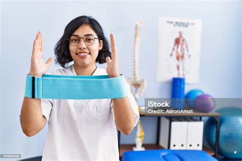 물리 치료 클리닉에서 탄성 밴드를 들고 물리 치료사 유니폼을 입은 젊은 라틴 여성 물리치료사에 대한 스톡 사진 및 기타 이미지 물리치료사 스트레칭 갈색 머리 Istock