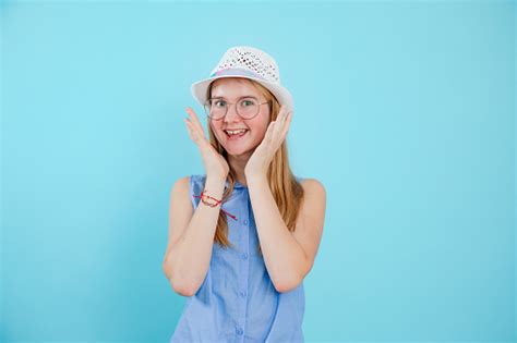 쾌활한 십대 소녀는 빈 파란색 배경에 행복한 감정으로 서 있습니다 세련된 민소매 셔츠 안경 및 귀여운 파나마를 입은 웃는 학령기 어린이의 초상화 십대 패션 여름 착용 감정에