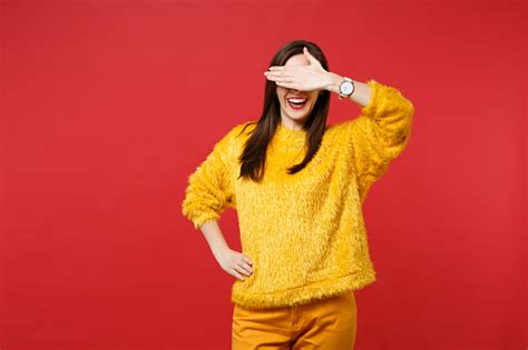 노란색 모피 스웨터 숨기기에 웃는 젊은 여자의 초상화 스튜디오에서 밝은 빨간색 벽 배경에 고립 된 손으로 눈을 덮고 사람들은 진심으로 감정 라이프 스타일 개념입니다 복사