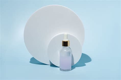 단단한 그림자가 있는 파란색 배경에 스포이드 뚜껑이 있는 유리병 모형 얼굴 혈청이 있는 컨테이너는 기하학적 둥근 연단에 가까이