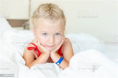 금발 머리를 가진 어린 소녀는 흰 리넨과 침대에 누워 있습니다 카메라에 미소를 짓고 보이는 아름 다운 아이의 초상화 빛 사진입니다 가리기에 대한 스톡 사진 및 기타 이미지