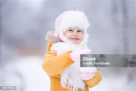 추운 날에 눈덩이를 만들고 들고 행복한 미소 짓는 2 세 유아 겨울 배경에 모피 모자 흰색 스카프 장갑과 노란색 따뜻한 재킷을 입은 한 아기 소녀 근접 촬영 전면 보기입니다
