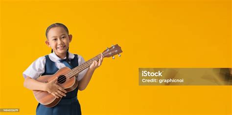 아시아 학생 귀여운 어린 소녀가 우쿨렐레를 연주하는 교복을 입고 디자인 작업을 위한 클리핑 경로가 있는 노란색 배경에 격리되어 있습니다 빈 여유 공간 콘서트에 대한 스톡