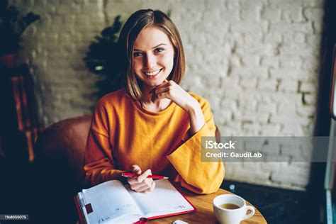 아침 시간에 커피 한 잔에 노트북에 메모를 만드는 카메라에 미소 세련된 짧은 헤어 스타일과 아름 다운 쾌활 한 학생의 절반 길이 초상화 긍정적 인 여성 소유자 쓰기 작업