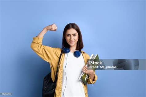 행운을 빌어 강한 웃는 학생 소녀는 근육을 보여주고 그녀의 지식에 자신있게 이정표를 통과하고 책을 들고 노란색 셔츠 흰색 티셔츠 검은 색 가방과 헤드폰을 목에 걸고 있습니다