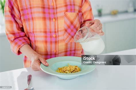 캐주얼 격자 무늬 셔츠를 입은 노인 여성은 아늑하고 햇볕이 잘 드는 부엌에서 콘플레이크 한 그릇에 우유를 부어 아침 식사를 준비하고 있습니다 50 54세에 대한 스톡 사진