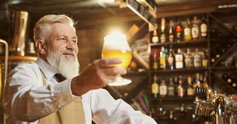 흰 셔츠와 앞치마를 입은 쾌활한 남자의 선택적 초점은 바 위에 서서 가벼운 맥주 한 잔을 유지합니다 머그잔으로 건배하는 동안 앞을 내다보고 웃는 남자 양조장의 개념입니다