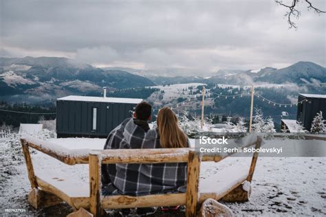 담요를 껴안고 겨울 산을 감상하는 사랑에 빠진 커플 레크리에이션 공간 현대적인 주택과 화덕 뒷마당의 벽난로 산 전망을 감상할 수 있는 통나무집 테라스 언덕 위의 초소형 주택