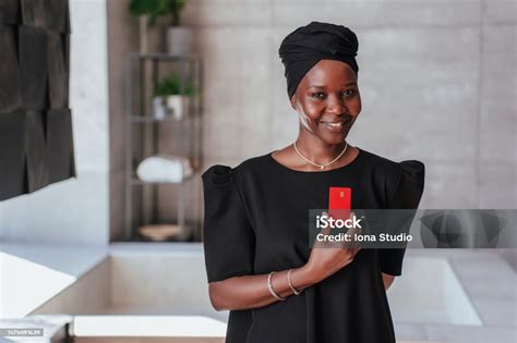 검은 터번과 검은 드레스를 입은 수줍은 아프리카 젊은 여성이 신용 카드를 들고 집에 서서 직업 경력 임금에 만족하며 카메라를 행복하게 바라보고 있습니다 성공한 사람들 재정적