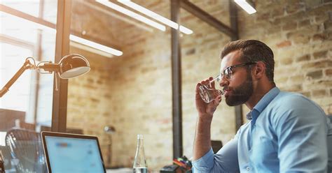 휴식이 필요해 안경을 쓰고 정장을 입은 목마른 수염 사업가는 현대적인 사무실에서 일하는 동안 신선한 물을 마시고 있습니다