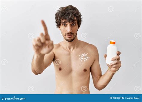 Young Hispanic Man Standing Shirtless Holding Sunscreen Lotion Pointing