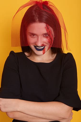 노란 스튜디오 배경에 고립 된 접힌 손으로 긴장한 포즈를 취하고 서있는 악마 여성의 정면보기는 빨간 베일과 검은 드레스를 입고 위험한 표정을 짓고 있습니다 할로윈 개념입니다