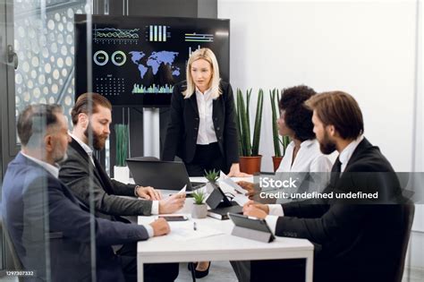 큰 Tv 플라즈마 스크린이있는 현대적인 회의실에서 다양한 다인종 동료들과 함께 회의에서 존경받는 집중 금발 여성 보스 리더가 세계지도 및 차트를 제시합니다 교육에 대한 스톡