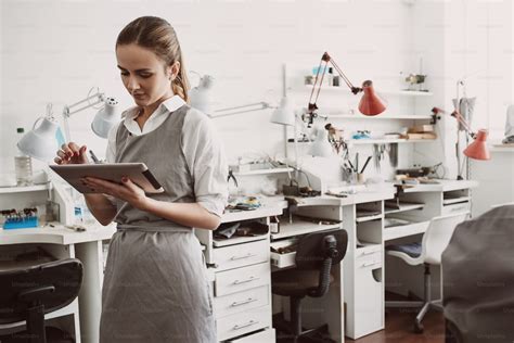 온라인 프로덕션 그녀의 보석 작업장에 서 있는 동안 그녀의 디지털 태블릿에 Crm 시스템을 사용하여 앞치마를 입은 젊은 여성 보석상의 초상화를 닫습니다 사업 보석