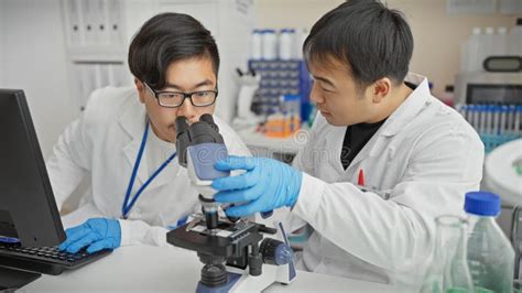 Two Men In Lab Coats Work Together In A Laboratory One Using A Microscope Highlighting