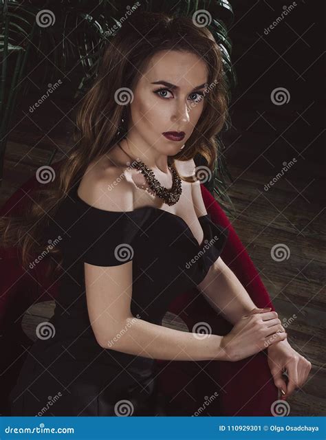 An Attractive Slender Slender Brown Haired Woman In A Long Black Evening Dress Sits On A Red