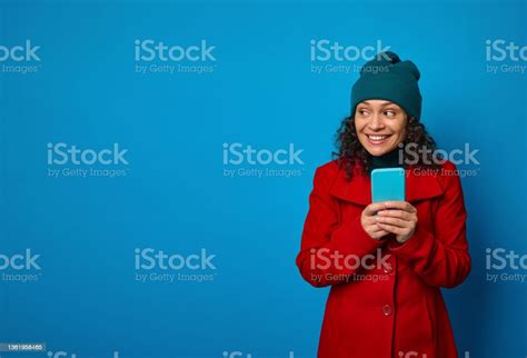 밝은 빨간 코트와 따뜻한 녹색 모직 모자를 쓰고 있는 쾌활한 행복한 예쁜 여성의 파란 배경에 고립된 초상화가 전화와 옆으로