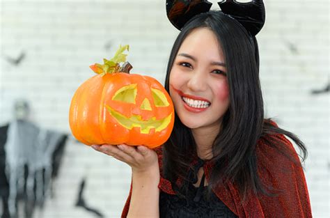 할로윈 파티를위한 검은 종이 박쥐 모양으로 장식 된 흰색 벽돌 배경에 호박 잭 O 랜턴을 들고 마녀 의상을 입고 젊은 아름다운 아시아 여성의 초상화 공휴일에 대한 스톡 사진