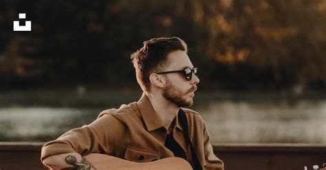 갈색 코트와 검은 바지를 입은 남자가 갈색 나무 의자에 앉아 갈색 어쿠스틱 기타를 연주하고 있다 사진 Unsplash의 무료 기타 이미지