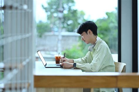Concentrated Young Asian Man Writing In A Notebook While Working Remotely With Laptop At Modern