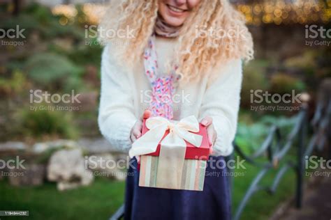 공원의 녹색 잔디 배경에 따뜻한 흰색 스웨터에 곱슬 금발 여자의 손에 새틴 리본과 함께 초점을 맞춘 선물 상자를 닫습니다 주는 과정을 선물합니다 소프트 선택적 초점 공간을