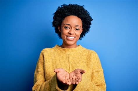 젊은 아름 다운 아프리카 계 미국인 Afro 여자 곱슬 머리 를 입고 노란색 캐주얼 스웨터 미소 와 함께 손바닥 을 받고 또는 제스처를 주는 보류 및 보호 손에 대한 스톡