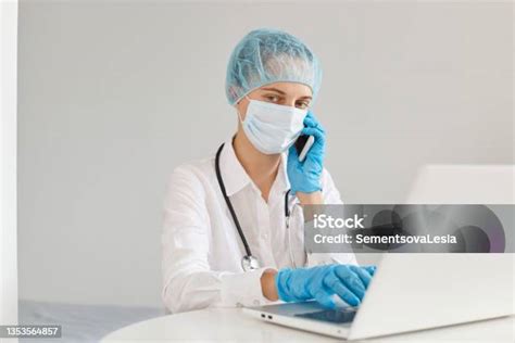 가운 수술용 마스크 의료용 모자 장갑을 착용한 진지한 여성 의사의 초상화가 노트북 앞에서 포즈를 취하고 대학과 전화통화를 하고 병원에서 일하고 있습니다 Brand Name