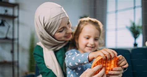 오렌지 안티 스트레스 감각 장난감 피젯 푸시 팝 히잡을 쓴 아름다운 무슬림 여성 집에서 소파에 앉아 그녀의 작은 귀여운 딸과 함께 다채로운 팝 그것을 안절부절 장난감으로