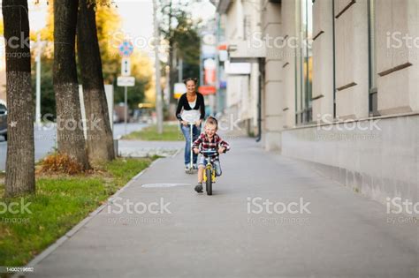 그녀의 아들과 함께 귀여운 젊은 어머니는 함께 도시의 보도를 타고 아이는 균형 자전거를 타고 엄마는 스쿠터를 타고 젊은 재미있는 엄마와 세련된 아이 25 29세에 대한 스톡