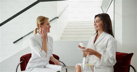 하얀 목욕 가운을 입은 매력적인 여자의 측면 모습은 뜨거운 음료수 한 잔을 들고 미용사와 이야기를 나눴다 그들은 편안한 의자에 앉아 웃고 있습니다 사진