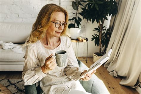 집에서 조용한 겨울 저녁 안락의자에 앉아 흥미로운 책과 차 잔을 손에 들고 둥근 안경을 쓰고 캐주얼하게 차려입고 거실 인테리어로 둘러싸인 예쁜 시니어 여성 여자에 대한 스톡