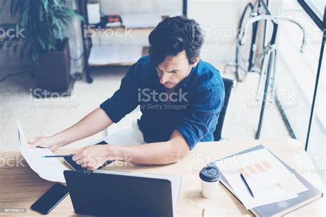 나무 테이블에 앉아 있는 동안 노트북에 밝은 직장에서 일 하는 젊은 동료 남자는 그의 손에 문서를 분석 합니다 흐리게 배경입니다