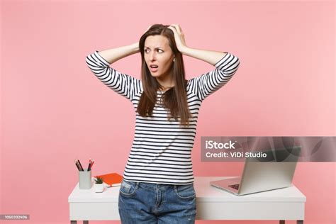 영 캐주얼 옷 파스텔 핑크 배경에 고립의 노트북과 흰색 책상 근처에 서 머리 작업에 매달리는 여자 우려 성과 비즈니스 경력