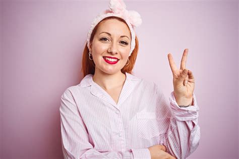 분홍색 고립 된 배경 위에 서있는 잠옷을 입은 젊은 아름다운 빨간 머리 여성은 승리의 표지판을하고있는 카메라에서 행복한 얼굴