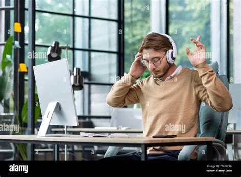 A Young Male Programmer Freelancer Sits In The Office At The Table With Headphones And Listens