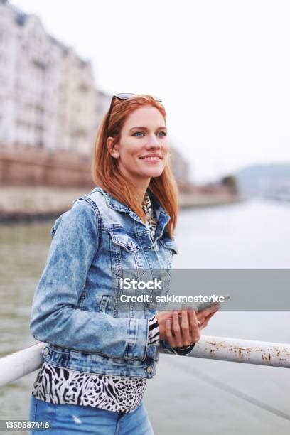 행복한 젊은 빨간 머리 여자 와 스마트 폰 에 강둑 멀리 보고 20 29세에 대한 스톡 사진 및 기타 이미지 20 29세 30 39세 Brand Name Online