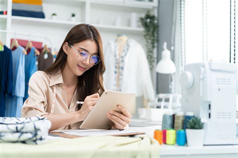 아시아 재단사 여성은 아틀리에를 재단사에서 새로운 옷을 디자인하기 위해 노력하고 있습니다 매력적인 젊은 여성 패션 디자이너 드레스 메이커는 태블릿을 사용하여 워크샵 룸에서