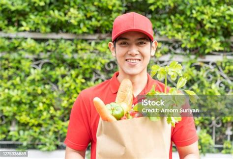 빨간 유니폼과 빨간 모자를 쓰고 미소 짓고 음식 과일 채소의 종이 봉투를 들고 있는 아시아 배달남자의 클로즈업 특급 배송 음식