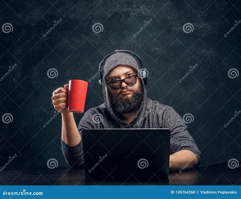 A Hacker Sits Behind A Laptop And Holding A Cup Of Coffee In A Dark Room And Stares Intently At