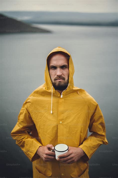 노란 비옷을 입은 잔인한 수염을 기른 남자가 금속 머그잔을 들고 호수의 전망을 감상하며 비오는 바람이 부는 날 절벽을 여행합니다