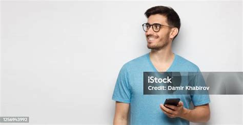 파란색 티셔츠와 안경을 쓰고 스마트폰을 손에 들고 서서 회색 배경에 고립된 공간을 복사하고자 하는 젊은 잘생긴 남자의 수평 배너 20 29세에 대한 스톡 사진 및 기타