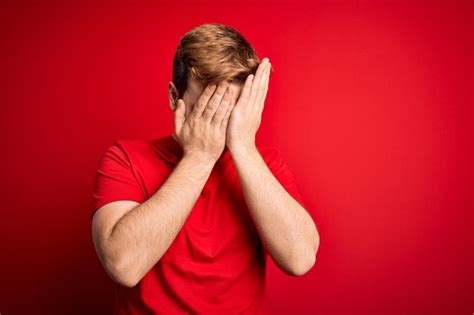 젊은 잘 생긴 빨간 머리 남자 는 울고있는 동안 손으로 얼굴을 덮고 슬픈 표정으로 고립 된 빨간 배경 위에 캐주얼 티셔츠를 입고 우울증 개념 수치심 부정적인 감정 표현에