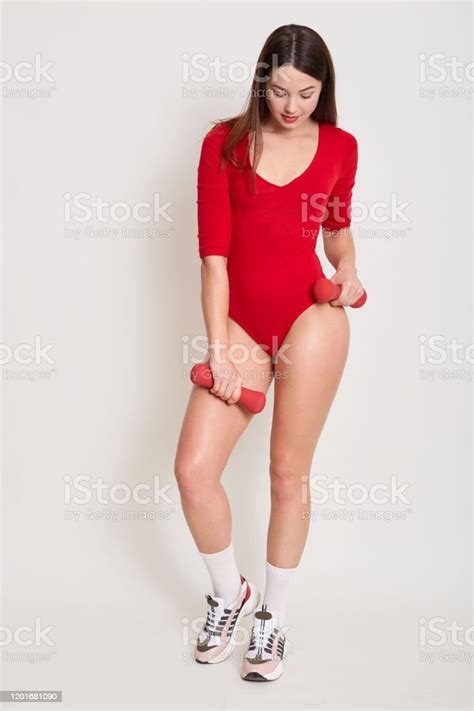 빨간 스포츠 콤비 드레스 흰색 양말과 운동화를 입고 아령을 들고 아래를 내려다 보며 스튜디오에서 흰색 배경 위에 고립 된 귀여운 아름다운 피트니스 트레이너의 전체 길이 사진