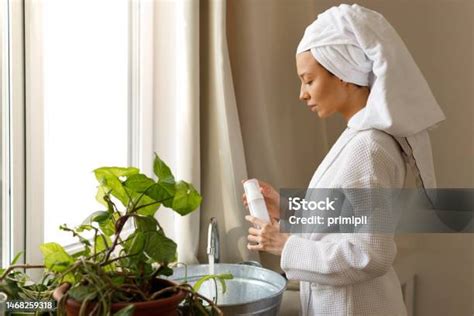 흰 가운을 입고 머리에 수건을 두른 예쁜 여자가 병에서 손바닥으로 젤이나 손 보습제를 붓습니다 샤워 후 건강한 바디 케어 화장실에 있는 소녀 샤워 젤에 대한 스톡 사진 및